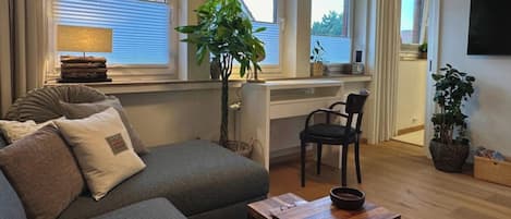 Uma sala de estar moderna com sofá, mesinha, cadeira e um espaço de trabalho com uma planta.