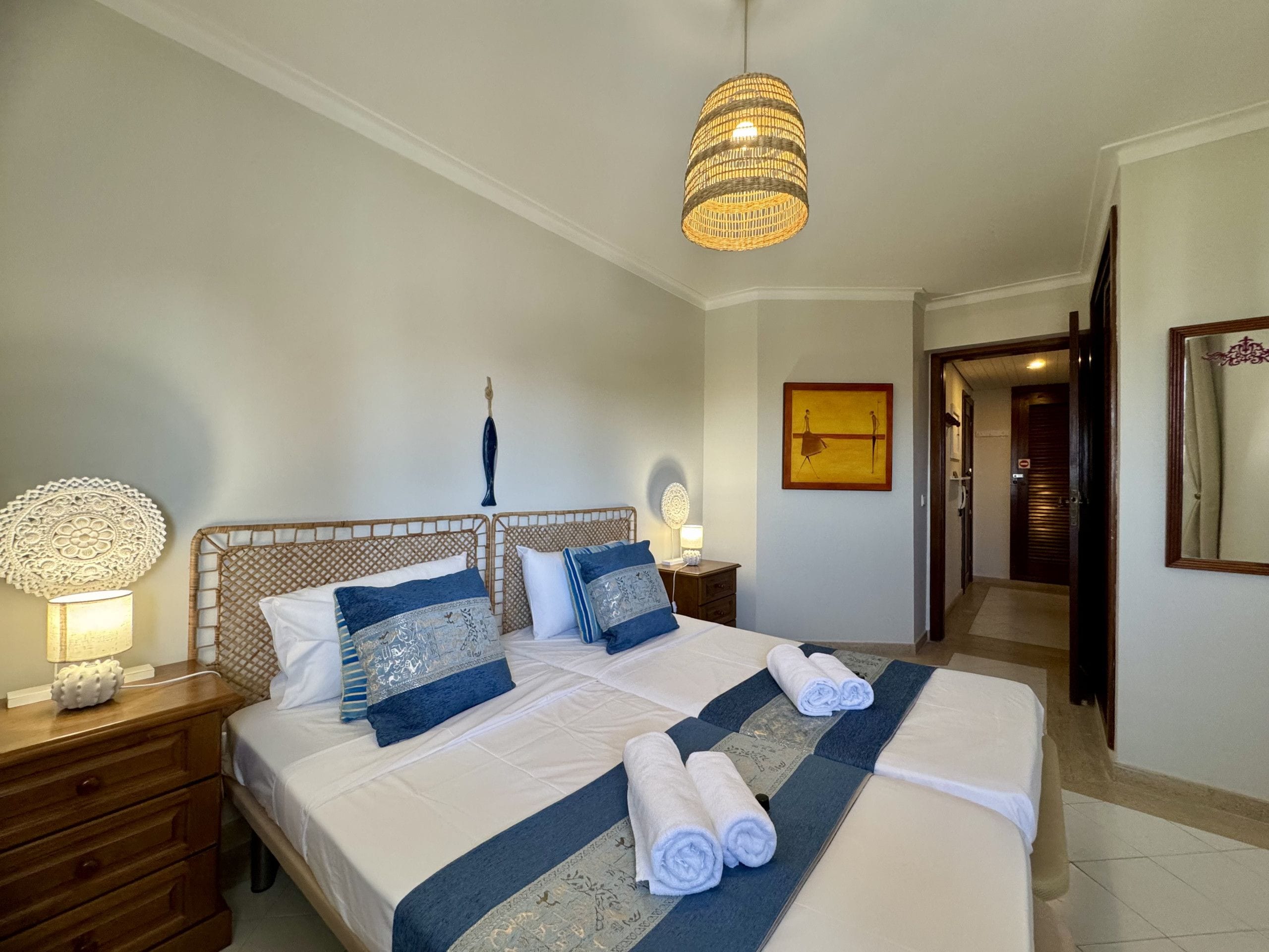 Quarto de hotel com uma cama grande, uma mesa de cabeceira com abajur, uma obra de arte na parede e uma porta que dá para outro quarto.