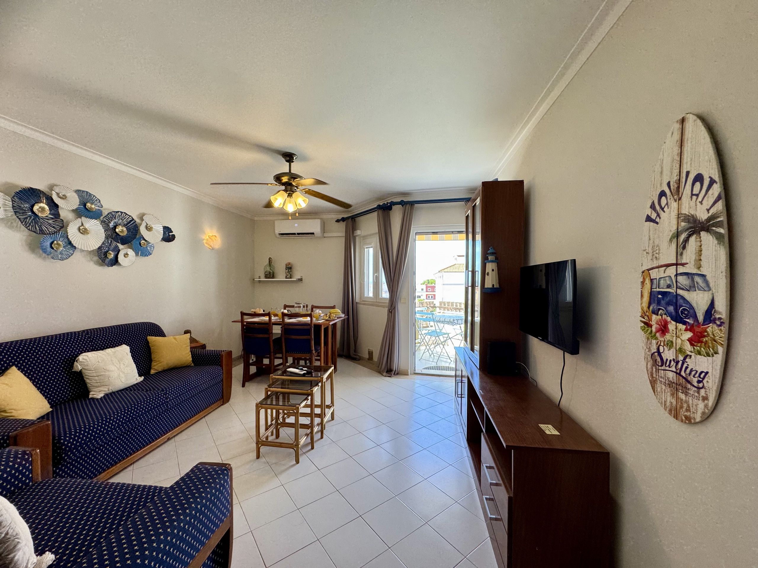 Uma sala de estar com sofá, mesa de jantar, cadeiras, ventilador de teto, televisão e uma decoração com prancha de surf.