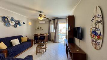 Uma sala de estar com sofá, mesa de jantar, cadeiras, ventilador de teto, televisão e uma decoração com prancha de surf.