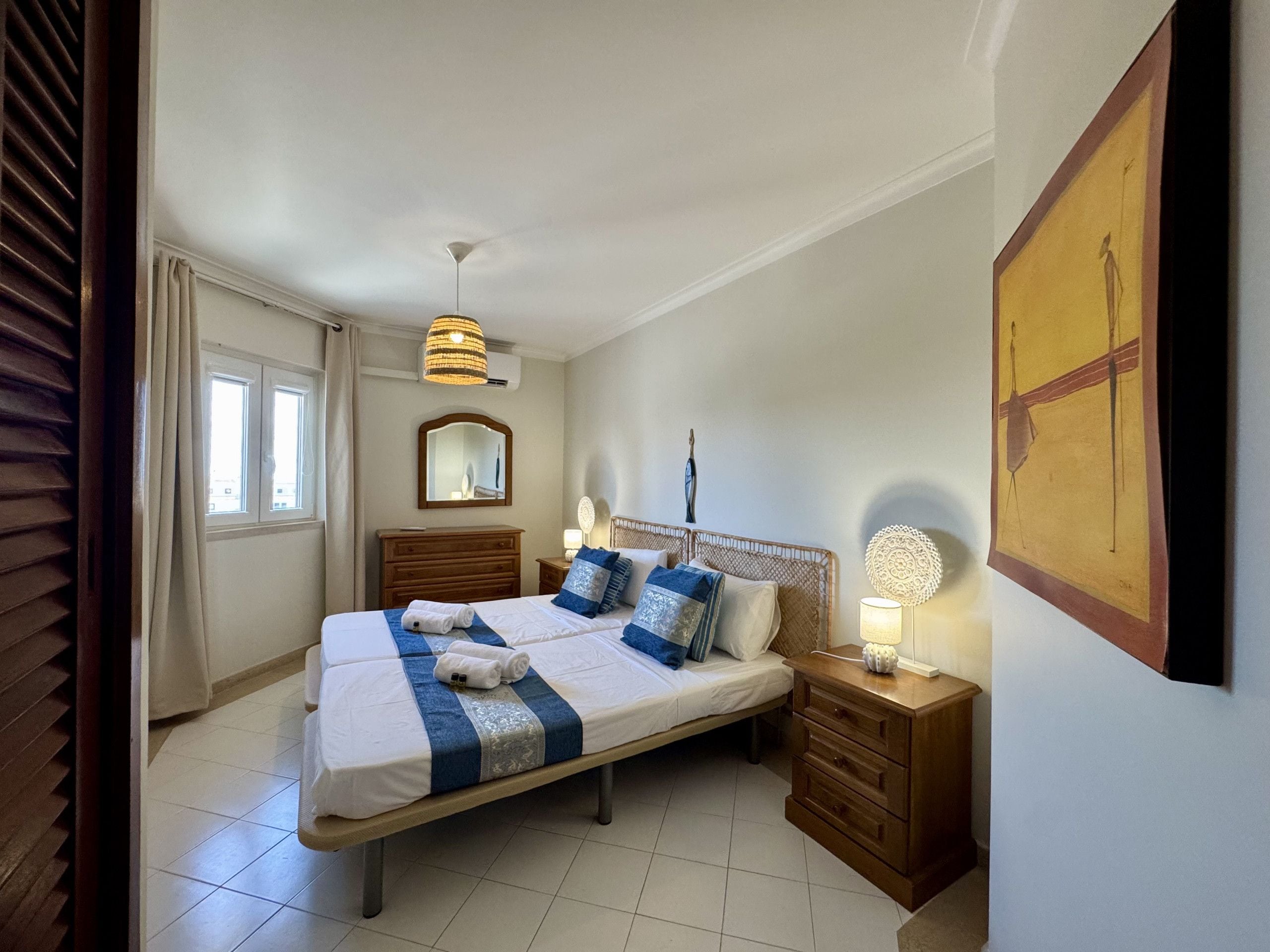 Quarto com cama, mesas de cabeceira, espelho, luminária pendente e uma obra de arte na parede.