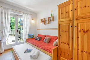 Quarto com cama, guarda-roupa de madeira, sofá com almofadas estampadas, pequena estante com objetos decorativos e vista através de uma porta de vidro.