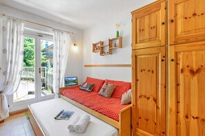 Quarto com cama, guarda-roupa de madeira, sofá com almofadas estampadas, pequena estante com objetos decorativos e vista através de uma porta de vidro.