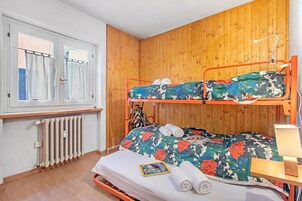 Uma cama beliche com colcha floral, uma janela com cortinas, um radiador e uma pequena mesa com um livro e toalhas.
