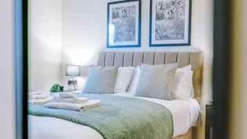 Cama bem arrumada com manta verde, almofadas brancas e cabeceira com duas fotografias emolduradas.