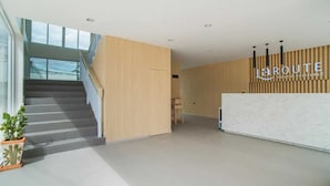 Uma área de recepção de hotel com uma escada que leva a um andar superior, um balcão de recepção e uma placa com a inscrição 'LAROUTE Hotel & Residence'.