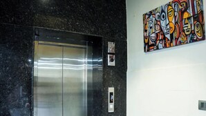 Um elevador em um hall de entrada com um visor digital, uma obra de arte na parede e uma câmera de segurança.