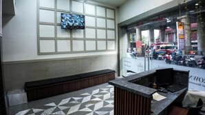 Uma área de recepção com piso de azulejos, um banco de espera, um balcão de atendimento e uma televisão fixada na parede.