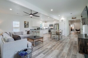 Uma sala de estar espaçosa com um sofá, mesa de centro e uma mesa de jantar com cadeiras. Uma cozinha com eletrodomésticos modernos pode ser vista ao fundo.