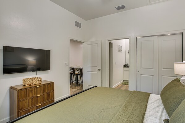 Quarto com cama, televisão na parede, penteadeira de madeira e porta que dá para outro espaço.