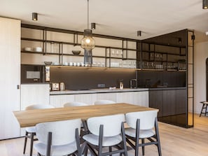 Cozinha moderna com mesa e cadeiras de madeira, estantes embutidas com copos de vidro e uma luminária pendente.