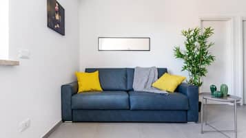 Uma sala de estar moderna com um sofá azul escuro, duas almofadas amarelas, um cobertor cinza, uma obra de arte emoldurada na parede, uma planta de bambu em vaso e uma pequena mesa com objetos decorativos.
