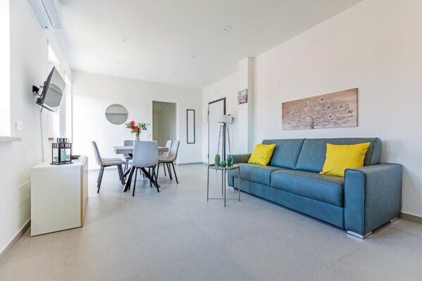 Uma sala de estar moderna com um sofá azul, um espaço de jantar com cadeiras, uma mesa de centro branca e uma TV de tela plana fixada na parede.