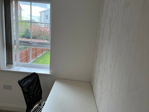 Quarto com escrivaninha branca, janela com vista para um jardim e um prédio, e radiador na parede.