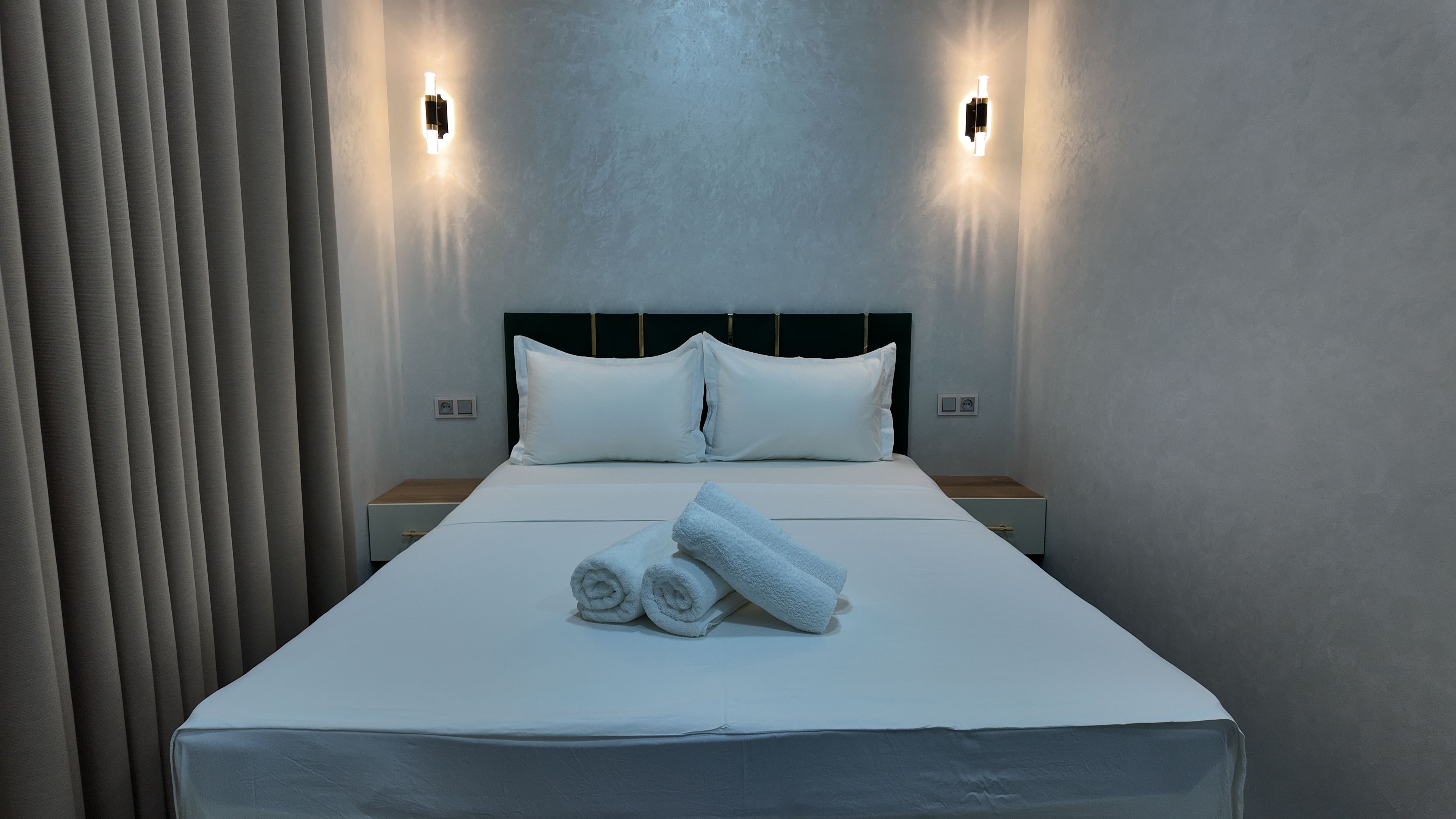 Quarto de hotel com uma cama grande, duas mesas de cabeceira e uma luminária fixada na parede.