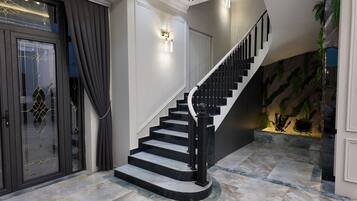 Escadaria com corrimão preto e degraus de mármore, que leva a um patamar com luminária fixada na parede.