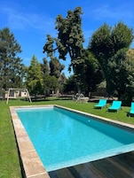 Piscina retangular com borda de deck de madeira, cercada por grama e árvores.
