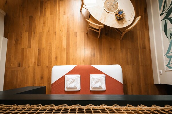 Quarto com piso de madeira, mesa redonda com cadeiras, cama com colcha vermelha e branca, e tapete de fibra natural.