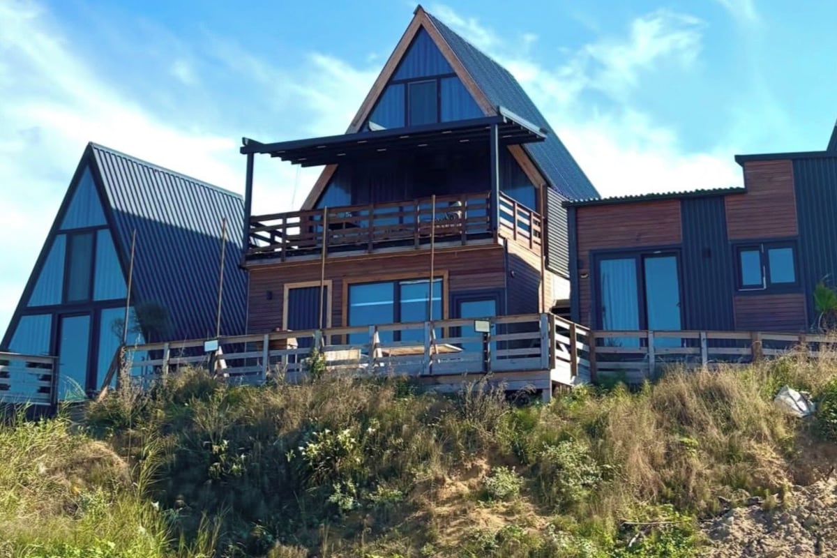 Uma cabana moderna de madeira, com vários níveis e uma varanda grande, com o céu azul claro ao fundo.