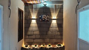 Um corredor estreito com parede de tijolos, duas luminárias fixadas na parede, uma obra de arte floral e um pequeno arranjo de flores no chão.