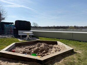 Uma área arenosa para brincar com um banco e uma mesa, cercada por uma cerca e um hedge.