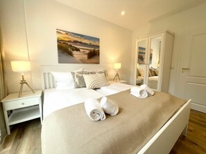 Cama bem arrumada com duas toalhas enroladas, abajur, mesa de cabeceira, espelho e um quadro de uma cena de praia na parede.