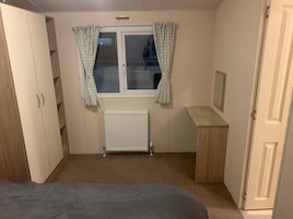 Quarto com cama, radiador, janela com cortinas, guarda-roupa e uma pequena mesa de madeira.