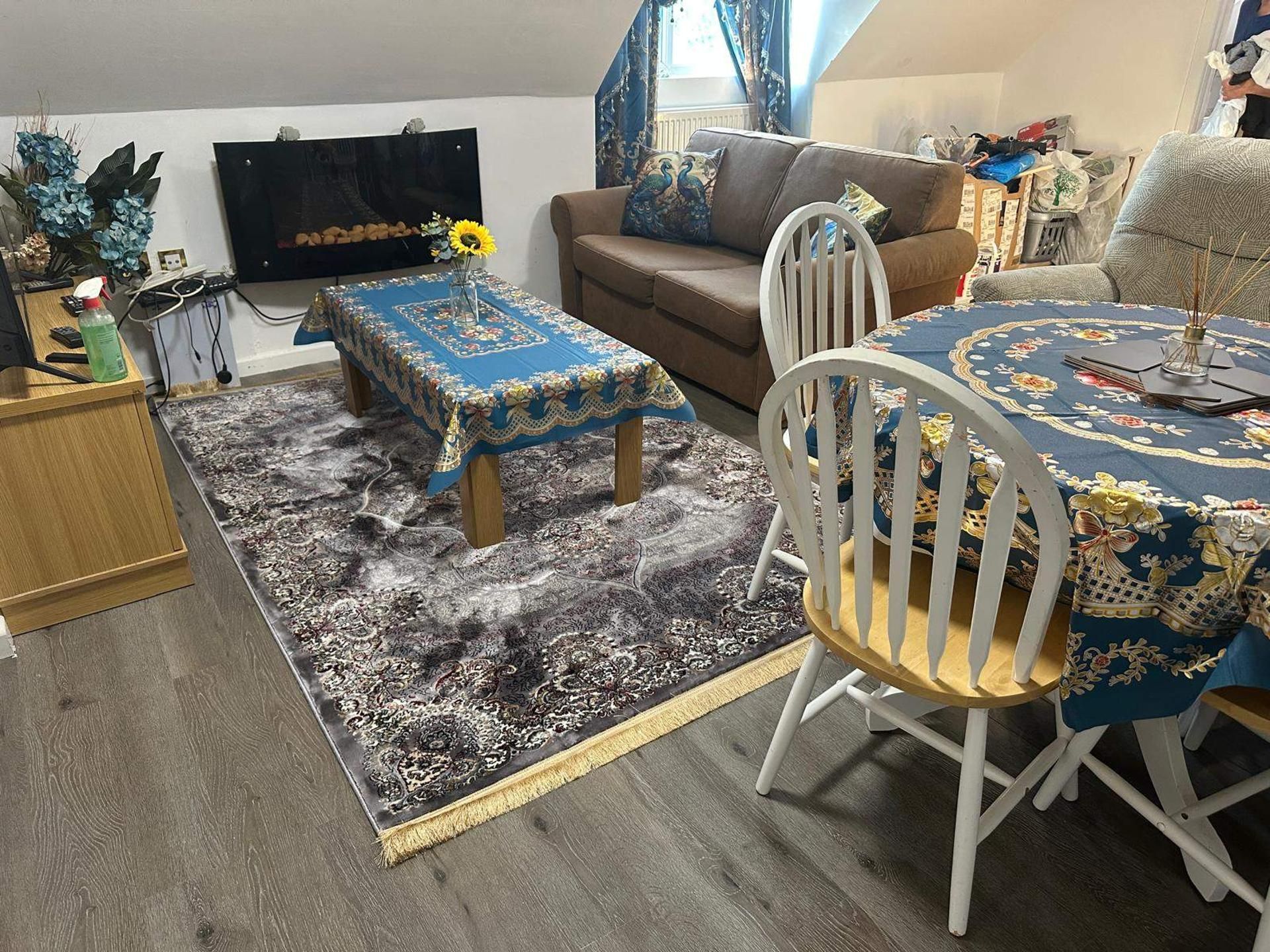 Uma sala de estar com sofá, mesa de jantar com cadeiras, televisão fixada na parede e tapete estampado.