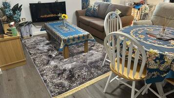 Uma sala de estar com sofá, mesa de jantar com cadeiras, televisão fixada na parede e tapete estampado.