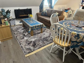 Uma sala de estar com sofá, mesa de jantar com cadeiras, televisão fixada na parede e tapete estampado.