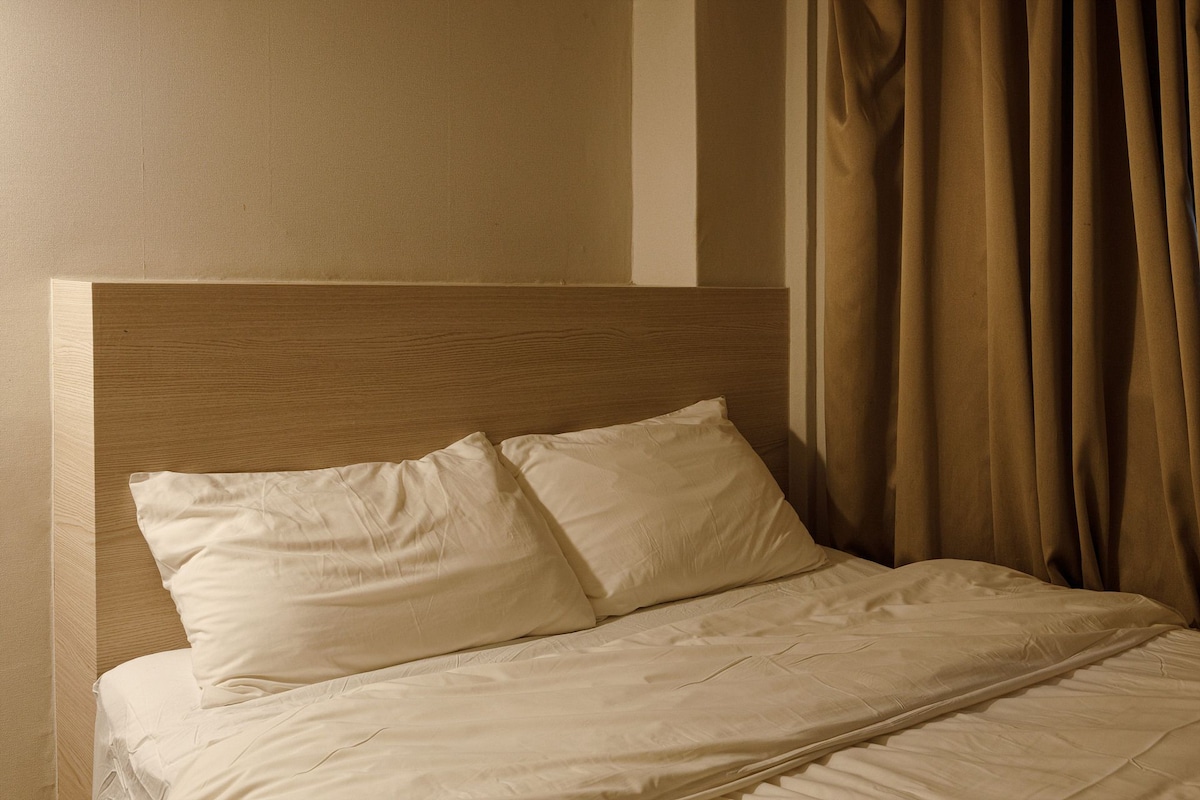 Cama com cabeceira de madeira e duas almofadas, em frente a uma parede lisa e uma cortina.
