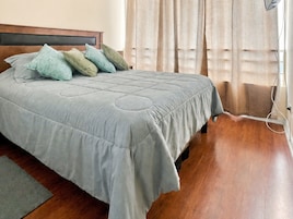 Cama bem arrumada com colcha cinza e várias almofadas, cabeceira de madeira e janela com cortinas.