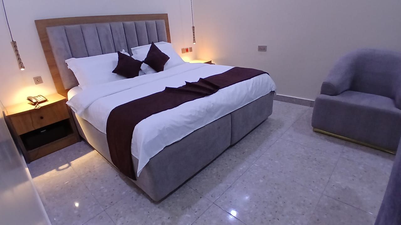 Uma cama bem arrumada com cabeceira, uma mesa de cabeceira com abajur e uma poltrona cinza.