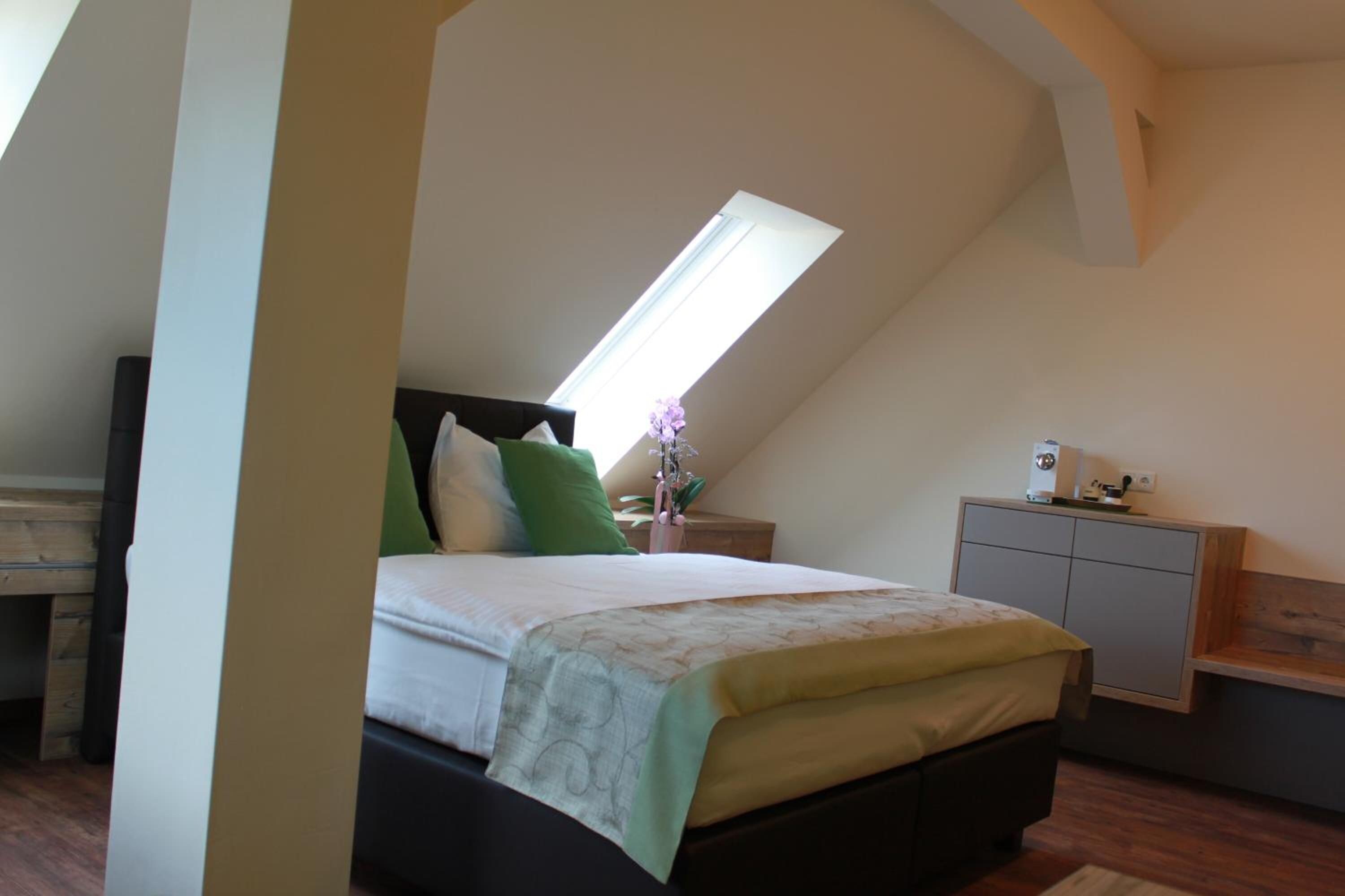 Quarto com teto inclinado, cama com colcha estampada, mesa de cabeceira com abajur e cômoda.