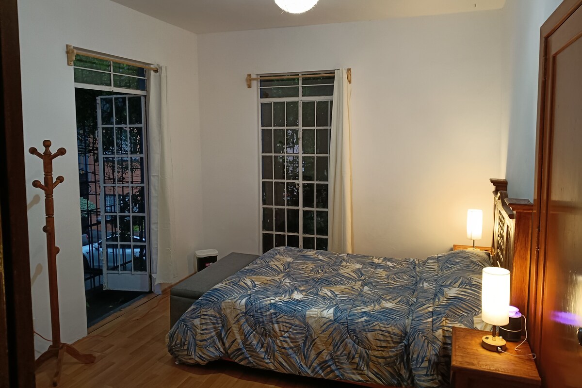 Quarto com cama, luminária de cabeceira, um banquinho de madeira, cabideiro e janela com cortinas.