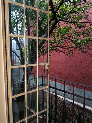 Janela com moldura de metal e maçaneta, com vista para uma parede vermelha e uma árvore com folhas verdes.