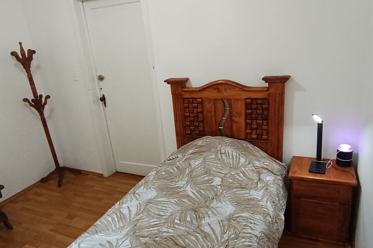 Quarto com cama de madeira, mesa de cabeceira com abajur e uma porta.