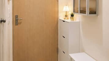 Um corredor com porta de madeira, um banco branco, uma mesinha com abajur e um armário com portas de vidro.