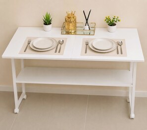 Mesa de jantar branca com dois jogos de pratos, talheres e guardanapos, adornada por pequenas plantas em vasos e um objeto dourado decorativo.