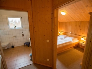 Interior de uma cabana de madeira com banheiro e quarto. O banheiro possui vaso sanitário, pia e uma janela com uma planta. O quarto tem uma cama, mesa de cabeceira e um abajur.