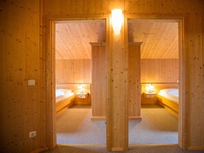 Quarto de madeira com duas camas, cada uma acompanhada de mesa de cabeceira e luminária, e chão coberto por carpete.