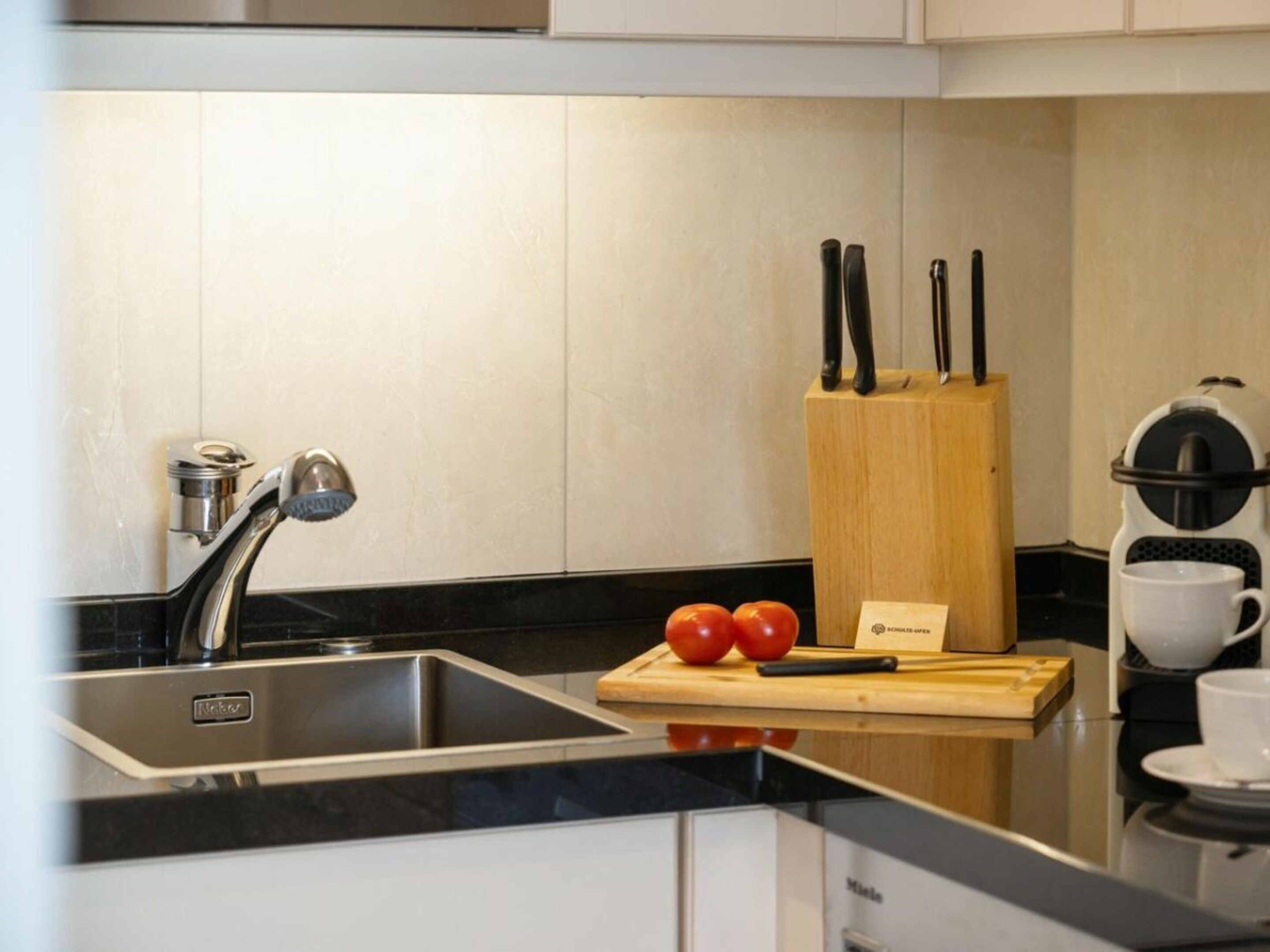 Uma cozinha moderna com uma pia de aço inoxidável, uma tábua de corte de madeira com tomates e facas, e uma cafeteira.