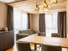 Uma zona de refeições moderna com mesa e cadeiras de madeira, área de cozinha com chaleira e grandes janelas com cortinas.