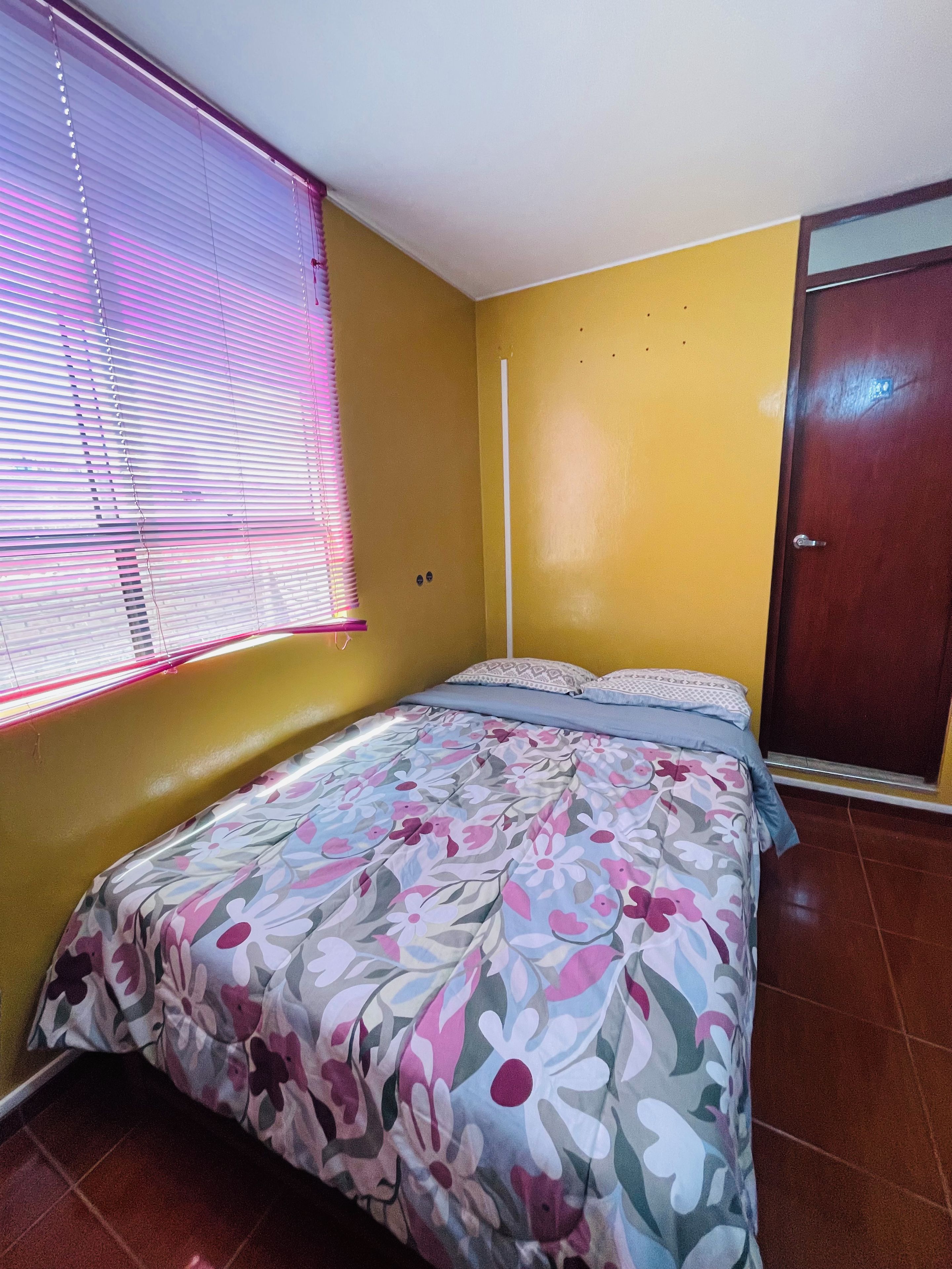 Quarto com cama, janela com persianas, porta e parede com interruptor de luz.