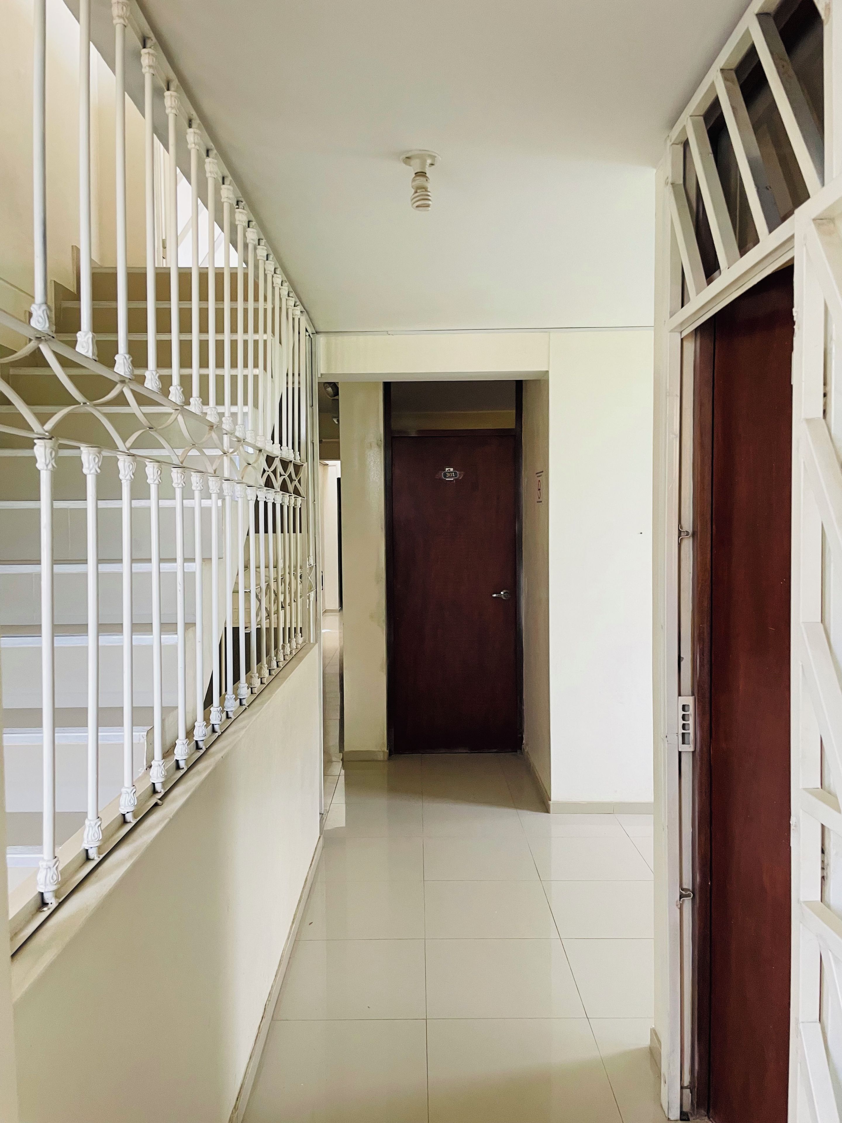 Um corredor com corrimão branco, porta de madeira e chão ladrilhado.
