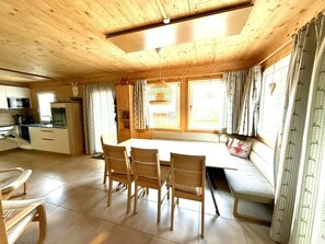 Interior em madeira com mesa de jantar, cadeiras, área de cozinha e janela com cortinas.