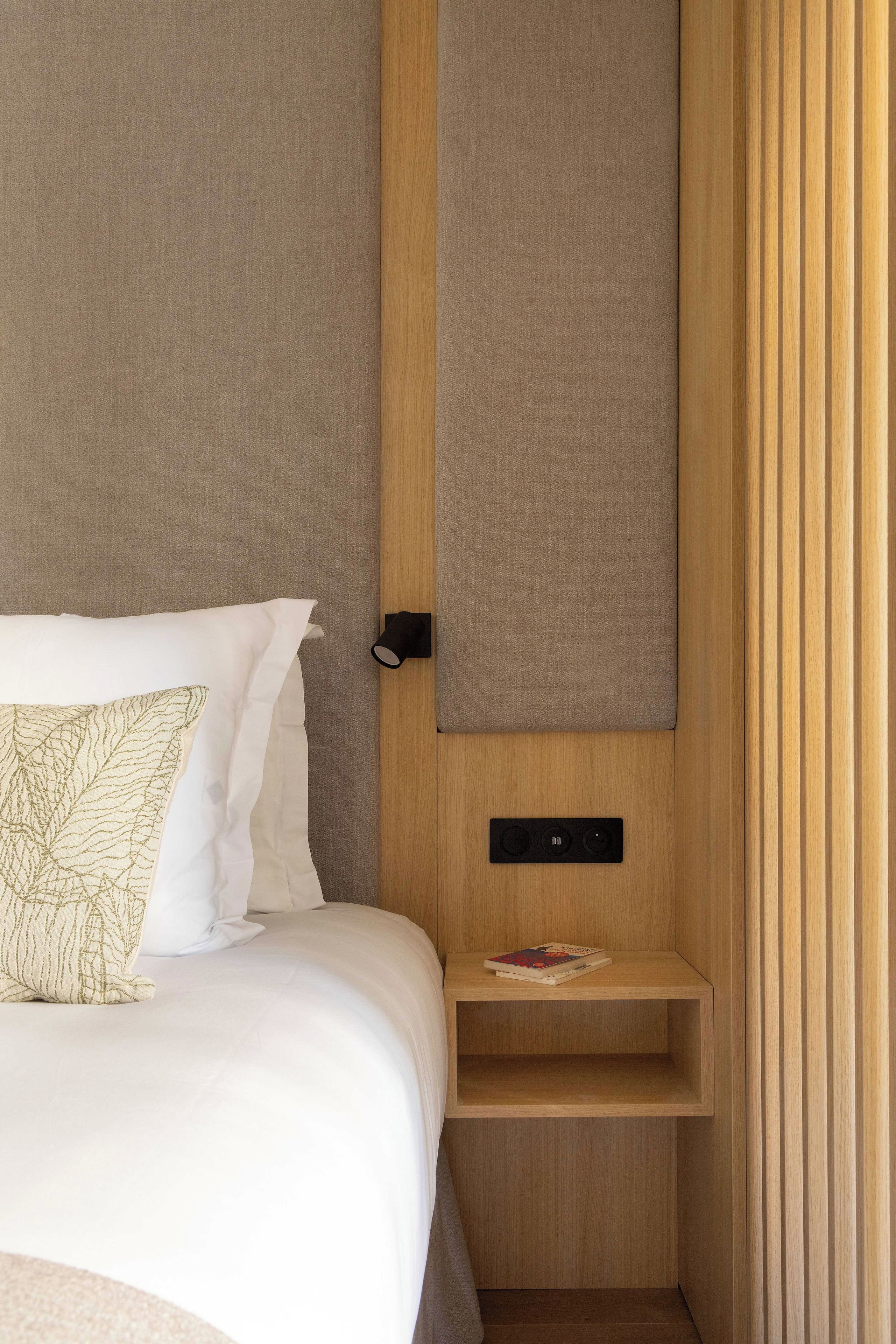Quarto de hotel com cama, mesa de cabeceira, luminária na parede e prateleira de madeira.