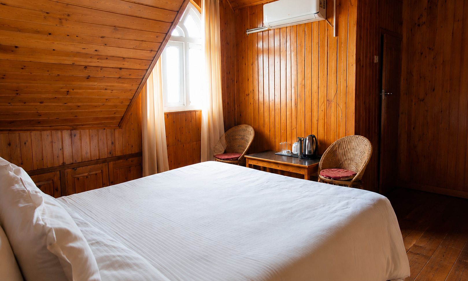 Quarto com cama, duas cadeiras de vime, uma mesinha com chaleira e uma janela.