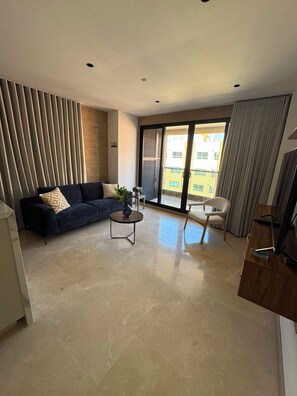 Uma sala de estar moderna com um sofá, uma mesa de centro e uma poltrona. Há uma janela grande com persianas e uma TV sobre um móvel de madeira.