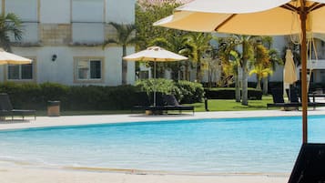 Área de piscina com espreguiçadeiras e guarda-sóis, um edifício com telhado em azulejos, e céu claro.